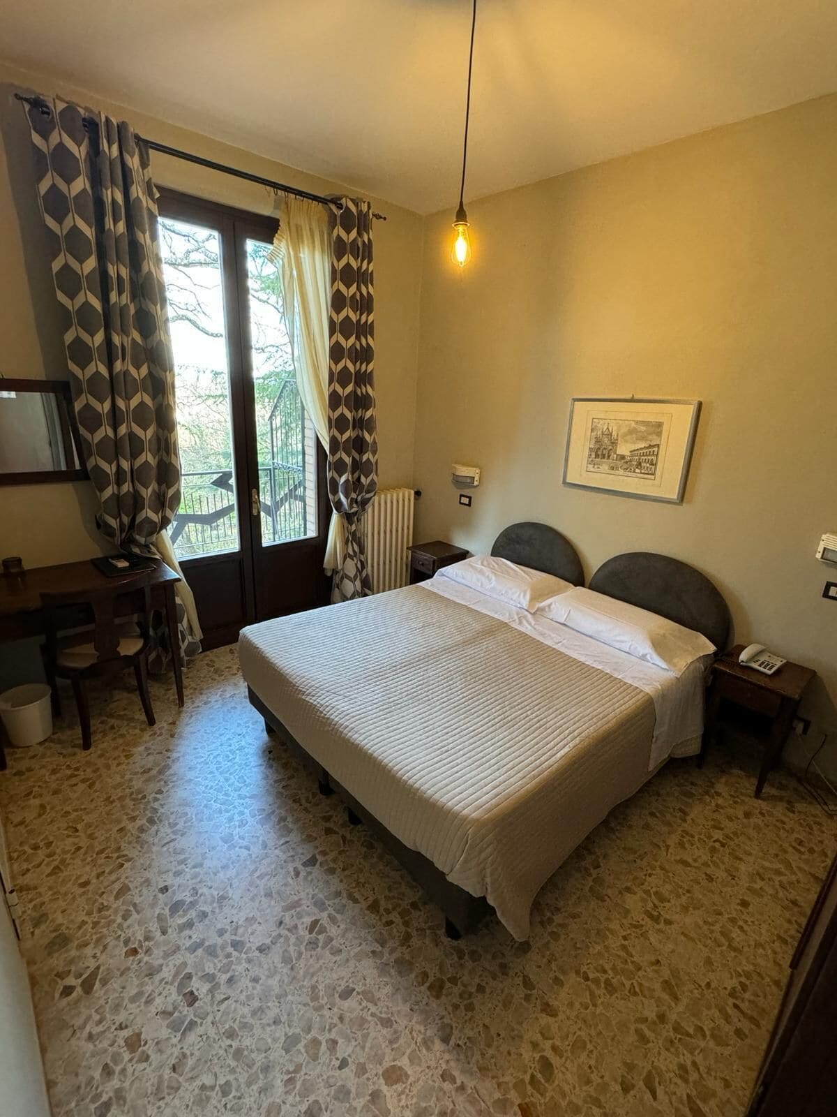Quarto com cama, escrivaninha, cadeira, radiador, telefone e um quadro na parede.