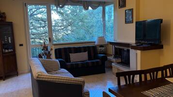 Sala de estar com sofá, televisão, mesa de jantar e janela com vista para árvores.