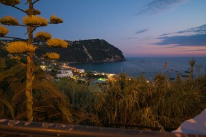 Uma vila costeira ao entardecer, com edifícios ao longo da orla marítima e uma colina imponente ao fundo.