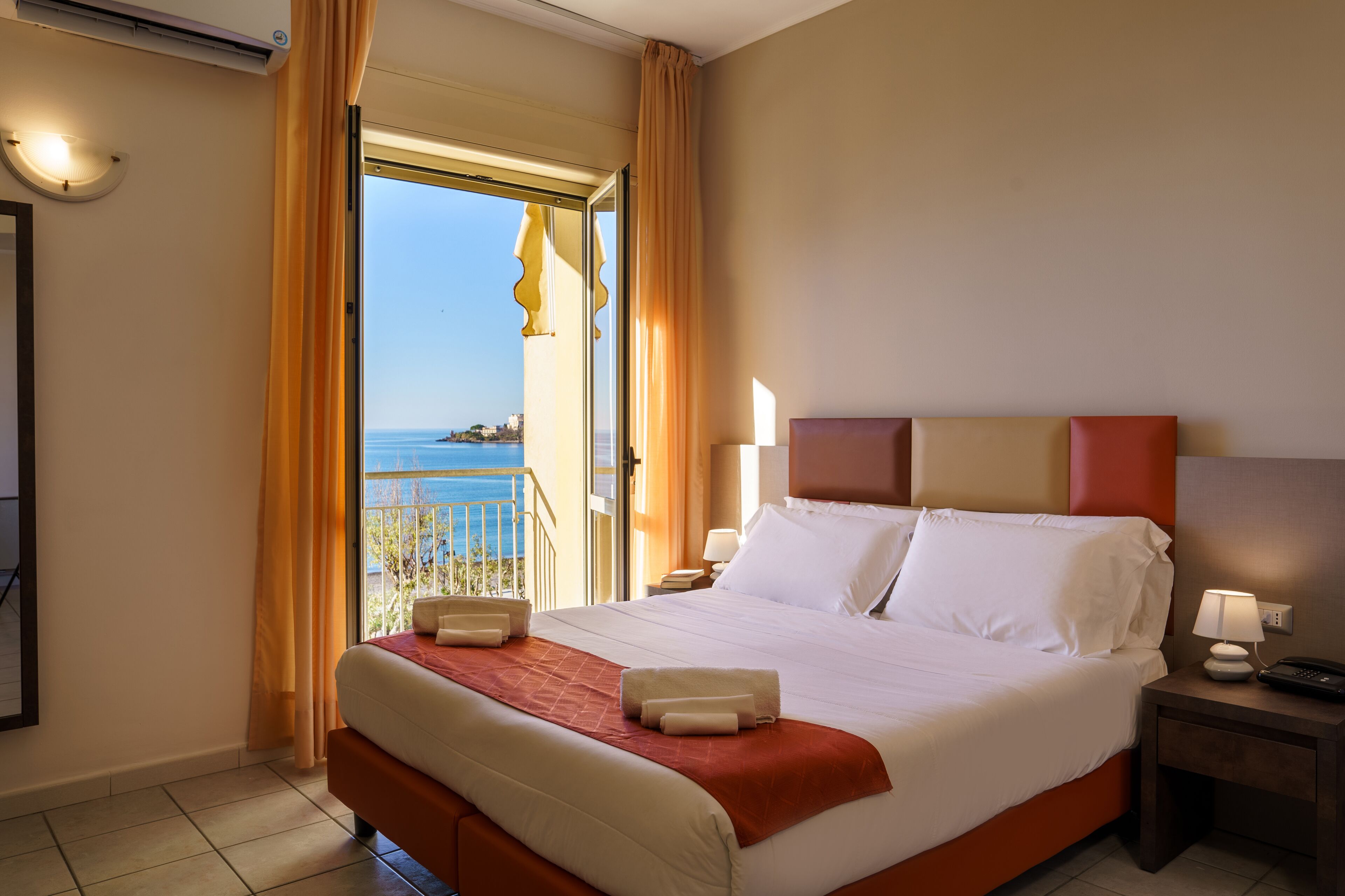 Quarto de hotel com uma cama grande, mesa de cabeceira com abajur e vista para o mar através de uma varanda.