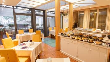 Restaurante com cadeiras e mesas amarelas, buffet com diversos pratos e grandes janelas que permitem entrada de luz natural.