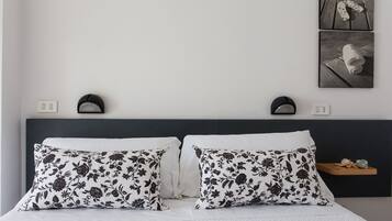 Cama bem arrumada com almofadas estampadas, cabeceira escura e prateleira de madeira pequena com objetos decorativos.