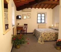 Um quarto pequeno e aconchegante com uma cama, uma escrivaninha e uma cadeira. Há uma janela com vista para o exterior e um vaso de planta no chão.