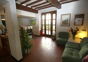 Sala de estar com sofá verde, mesa pequena com abajur e planta em vaso. Há uma porta que dá para uma área de cozinha com janela.