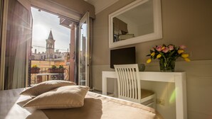 Quarto de hotel com cama, escrivaninha, cadeira, espelho e vista para um edifício com torre.