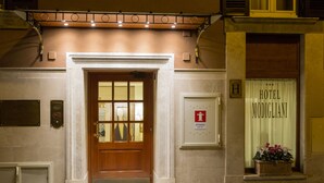 Entrada de hotel com porta de madeira, placa indicando saída de incêndio e floreira externa.