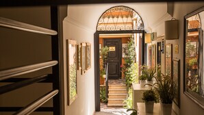 Um corredor com uma escada que leva a uma porta escura com a inscrição "The Apartment". Há plantas em vasos no lado direito e quadros nas paredes.