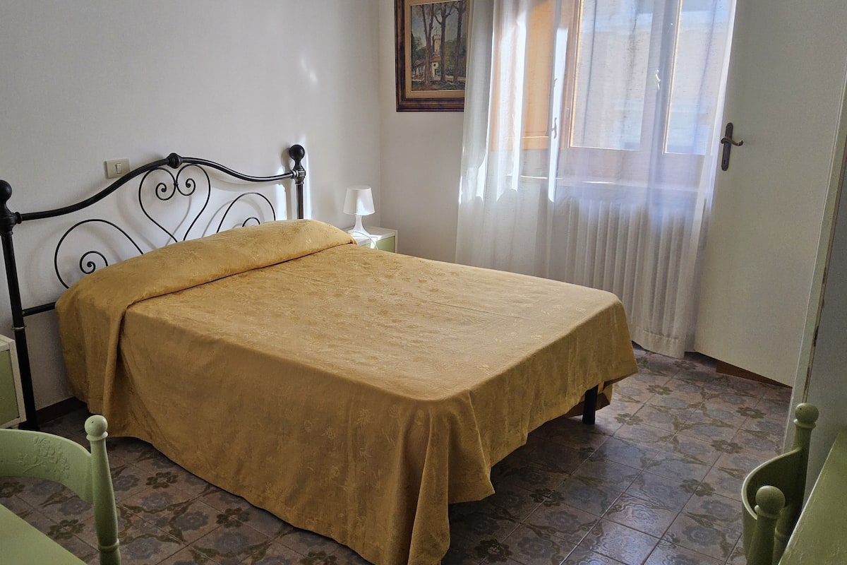 Quarto com cama, mesa de cabeceira, luminária, cadeira e janela com cortinas.