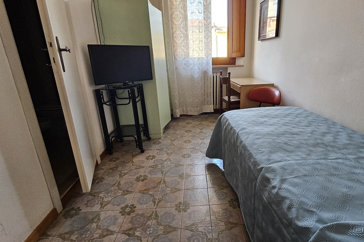 Um quarto pequeno e estreito, com uma cama, televisão em móvel, uma cadeira e uma escrivaninha.