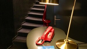 Quarto de hotel com escrivaninha, telefone vermelho, abajur e folheto do hotel.