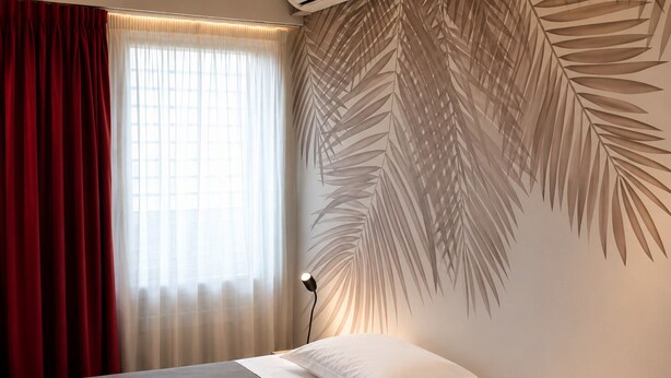 Quarto de hotel com uma cama, ar condicionado fixado na parede, janela com cortinas leves e parede com padrão de folha de palmeira.