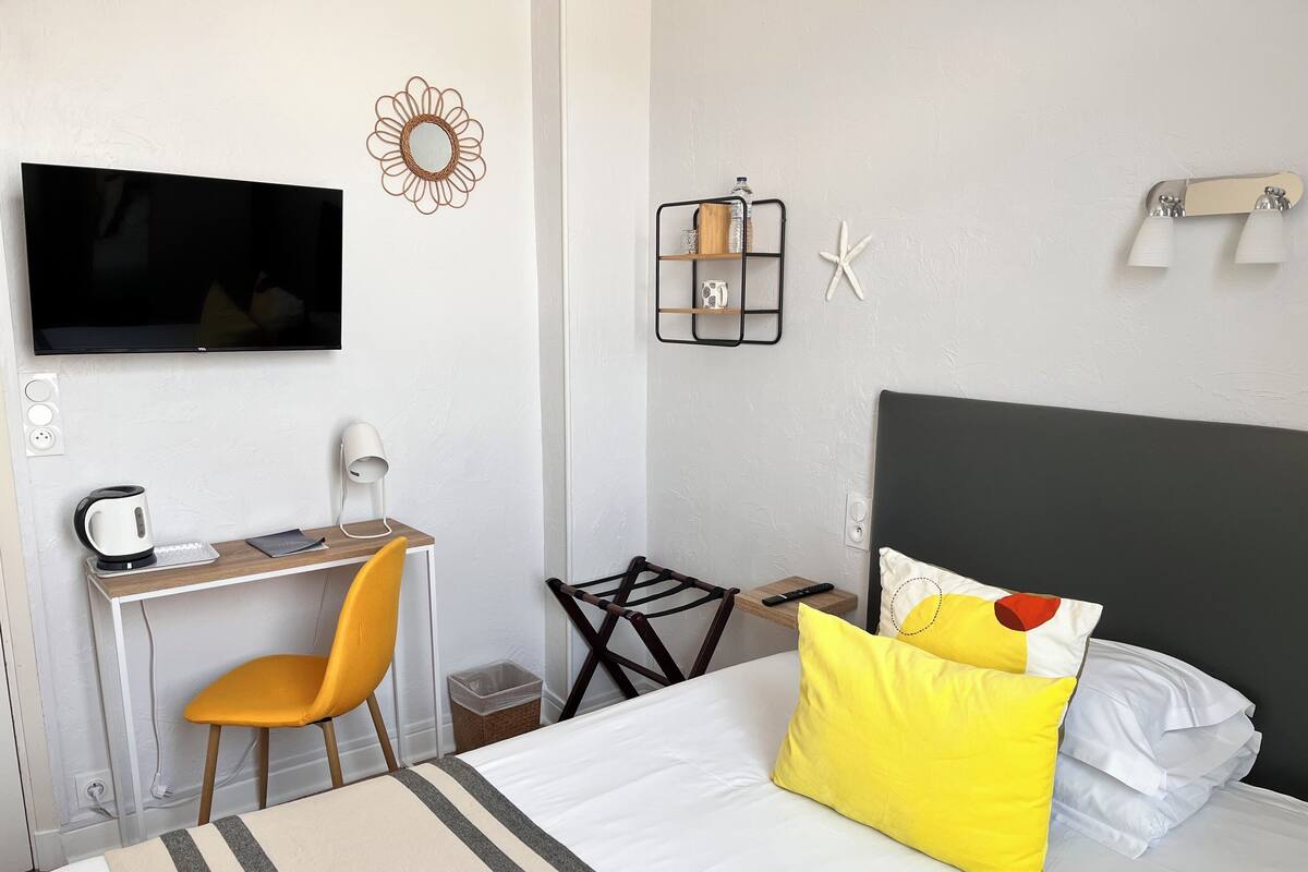 Quarto de hotel com cama, escrivaninha e cadeira, televisão, estante com objetos decorativos e mesa de cabeceira com abajur.