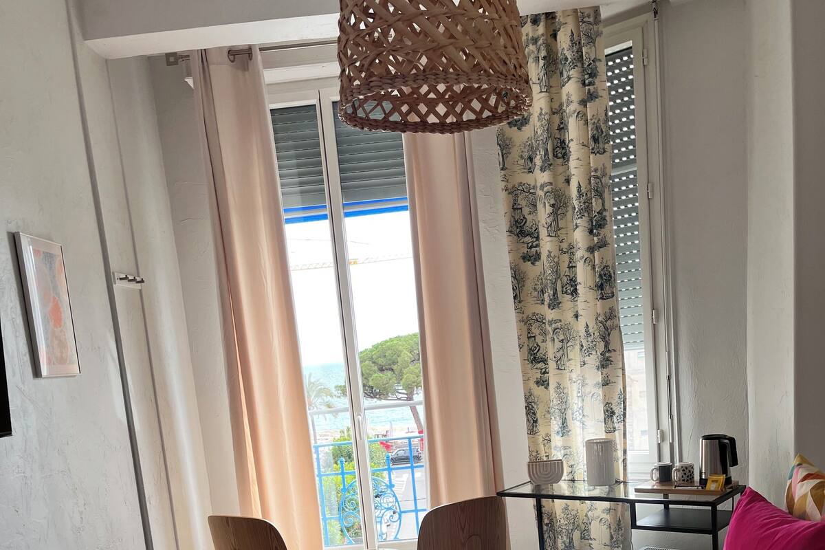 Quarto de hotel com uma cama grande, uma mesinha com cafeteira, duas cadeiras e vista para o mar pelas janelas.