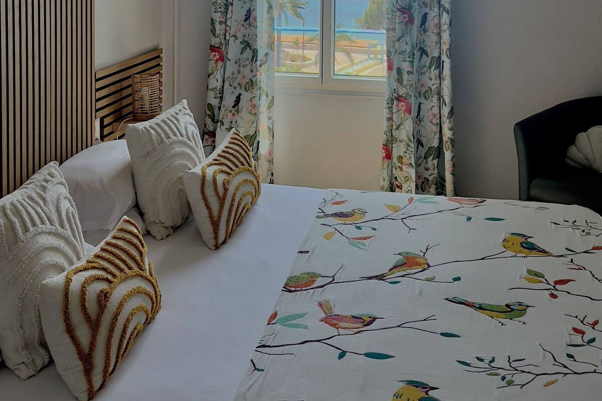 Quarto com cama, almofadas, luminária pendente, janela com cortinas e decoração na parede.