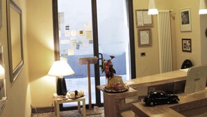Quarto de hotel com soalho em azulejos, uma escrivaninha com candeeiro, radiador e janela com vista.