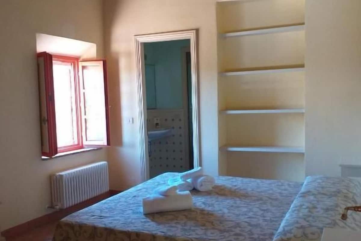 Quarto com cama, radiador, janela com molduras vermelhas e porta que dá para outro cômodo.
