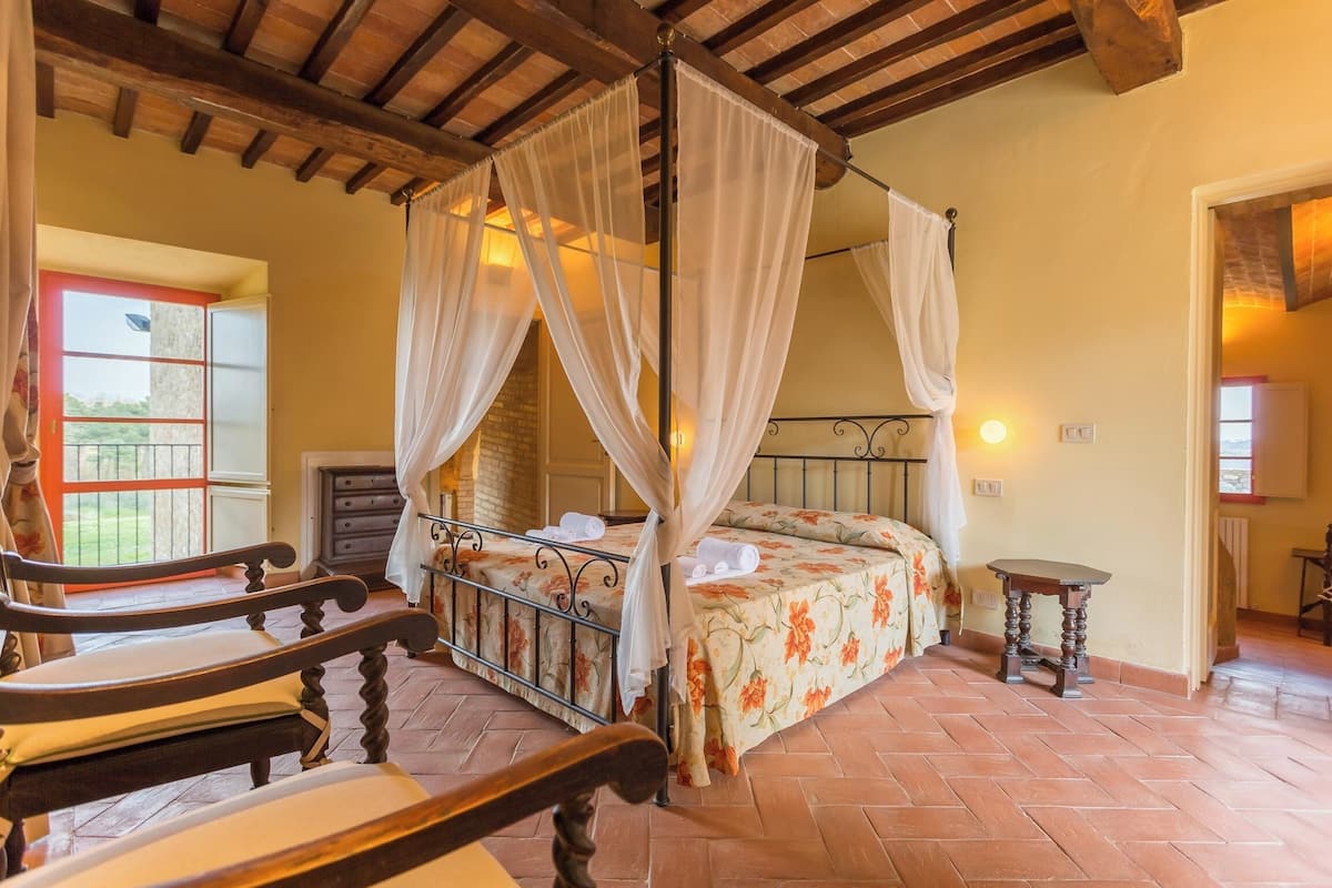 Quarto com uma cama de dossel, uma cadeira de madeira, uma mesinha e uma janela com cortinas.