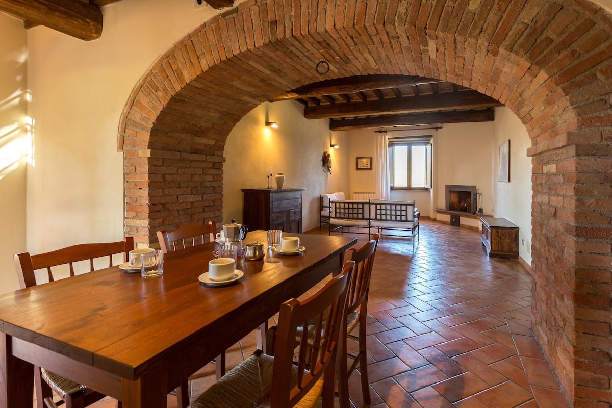 Uma sala de jantar com arco de tijolos, mesa de madeira, cadeiras e lareira ao fundo.