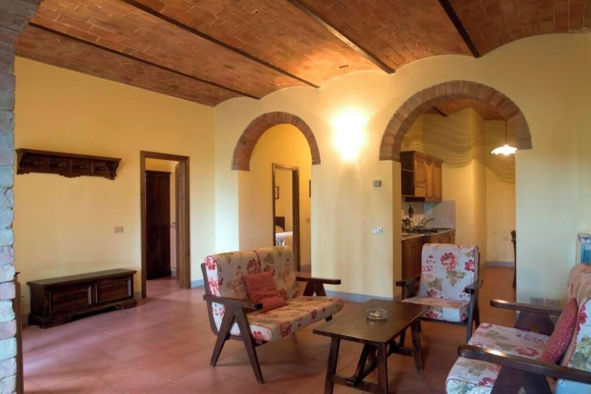 Uma sala ampla com teto alto, vigas de madeira e uma cozinha visível através de um arco.