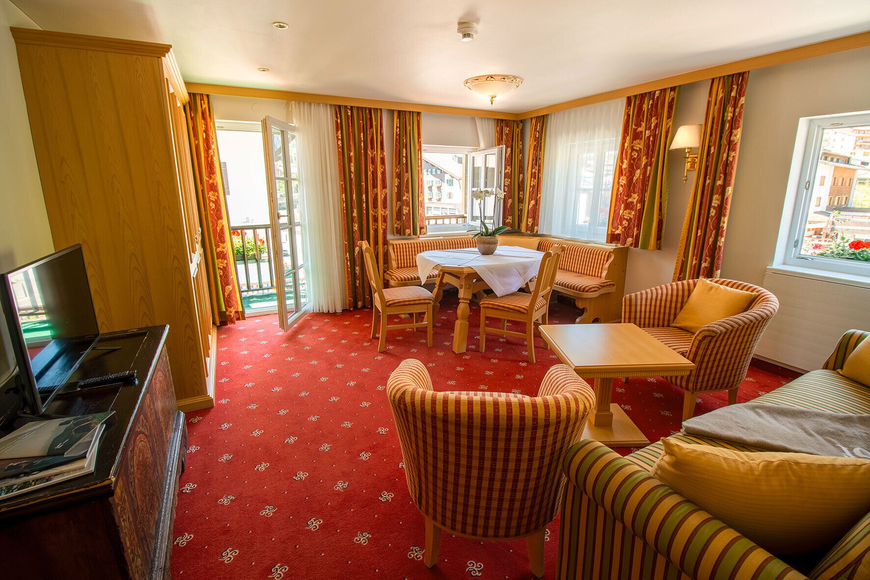 Quarto de hotel com carpete vermelho, poltronas listradas, mesa de jantar e televisão sobre um móvel de madeira.