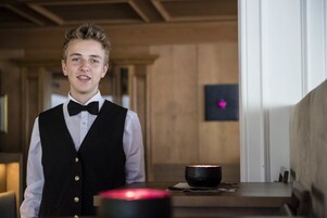 Garçom com colete preto e gravata borboleta em restaurante com paredes de madeira e design de interiores moderno.
