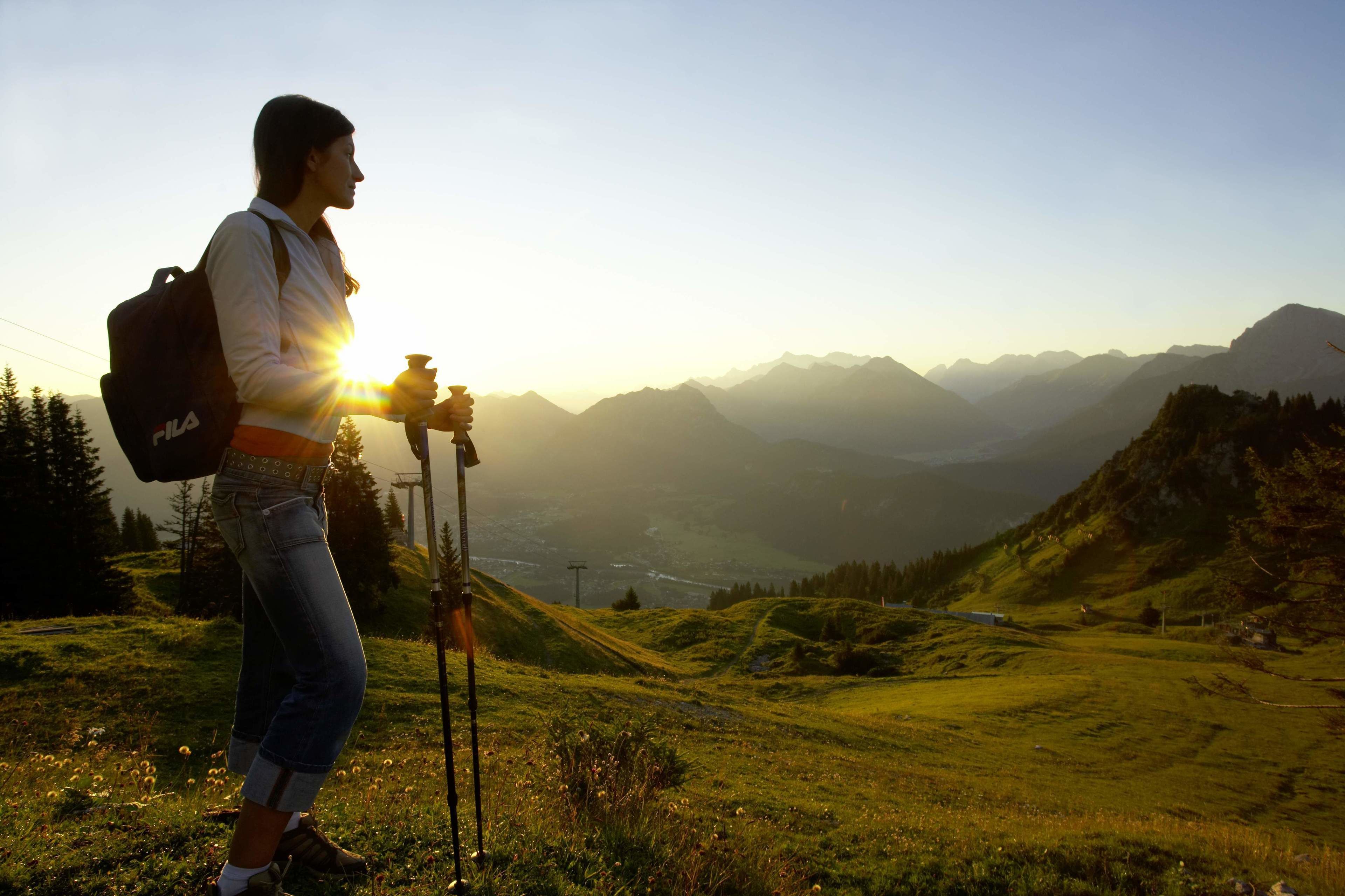 Uma pessoa caminhando com uma mochila e bastões de caminhada em uma encosta gramada, com uma cadeia de montanhas ao fundo.