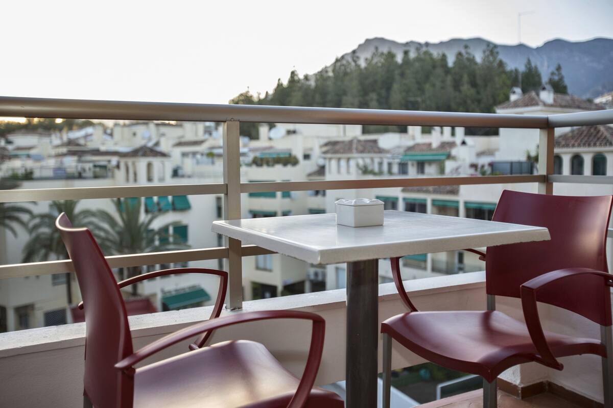 Uma varanda com mesa e duas cadeiras, com vista para um horizonte urbano com edifícios e montanhas.