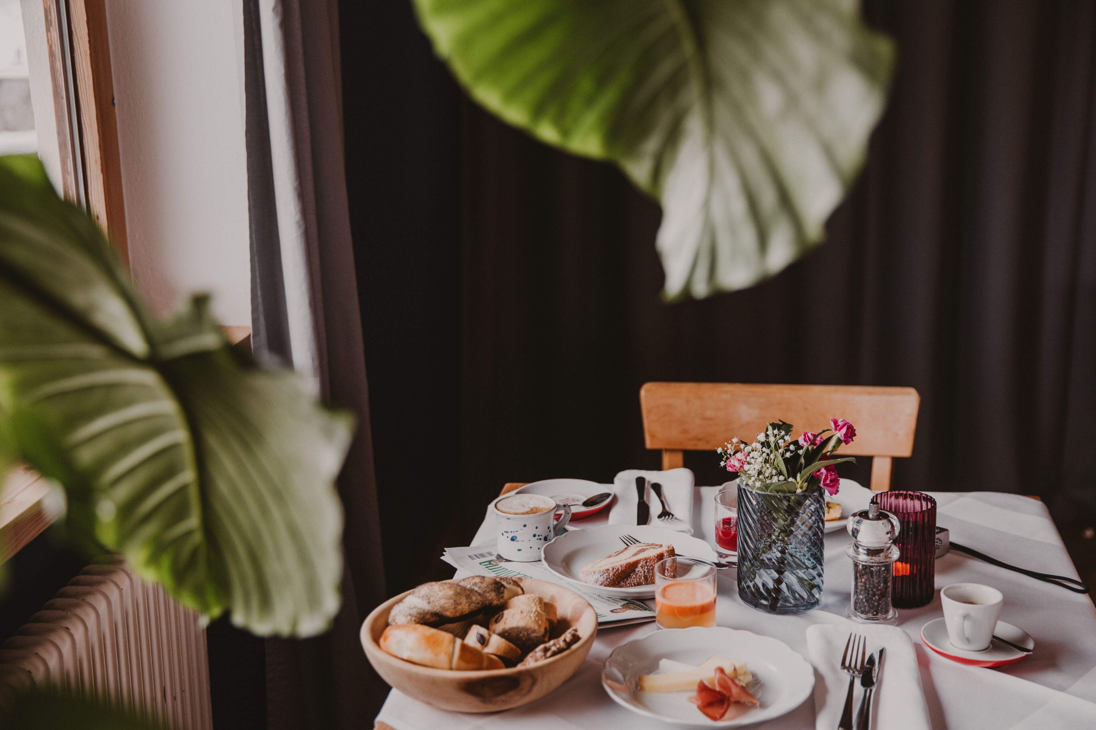 Mesa de madeira posta para o café da manhã, com pão em uma tigela, prato de doces, xícara de café e um vaso com flores.