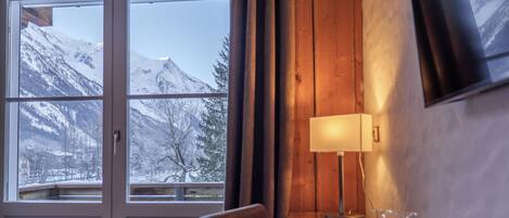 Quarto com uma grande janela que oferece vista para montanhas cobertas de neve. Há uma cadeira de madeira e uma mesa com um abajur.