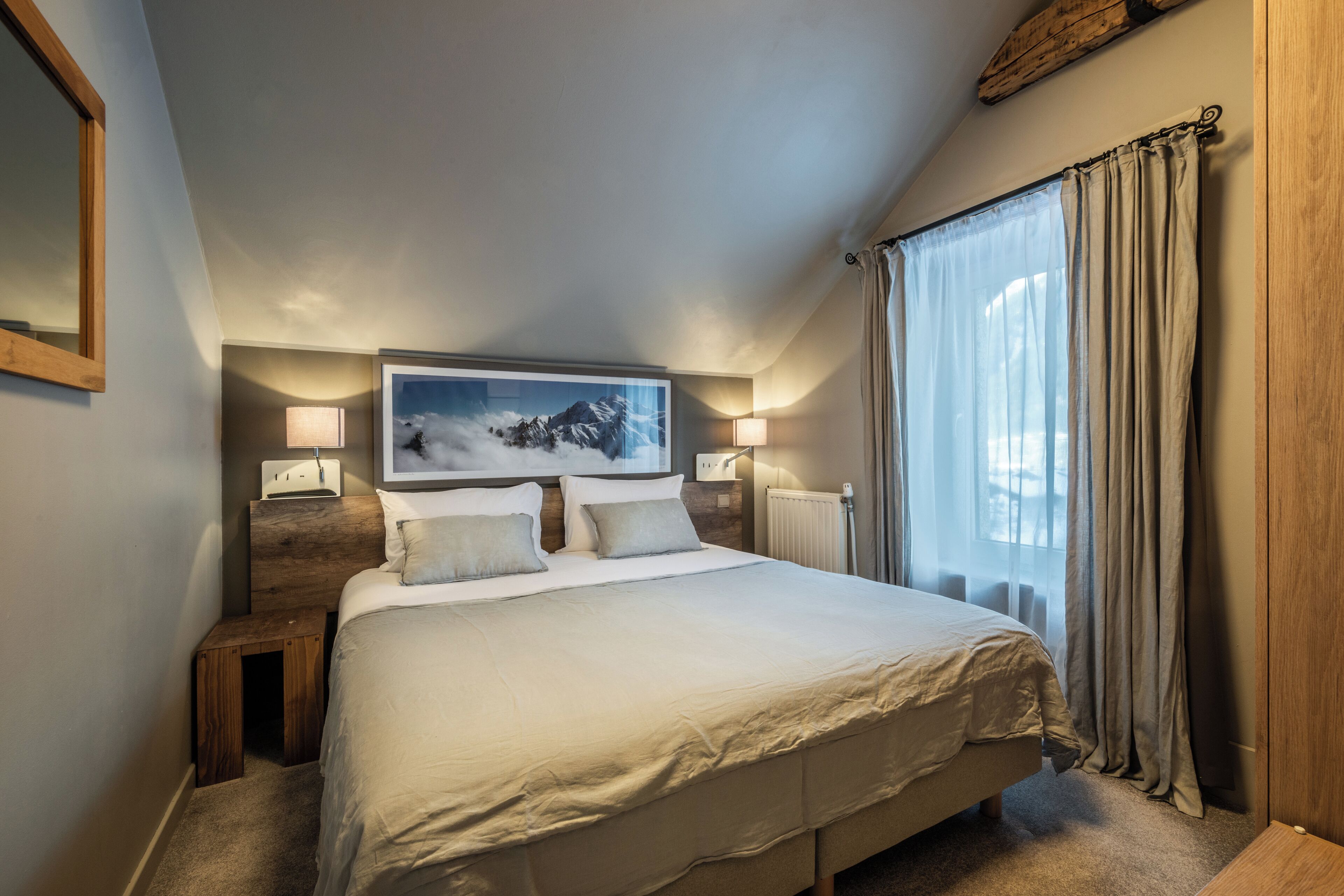 Quarto com uma cama grande, duas luminárias de cabeceira, uma mesa de cabeceira, um quadro na parede e uma janela com cortinas.