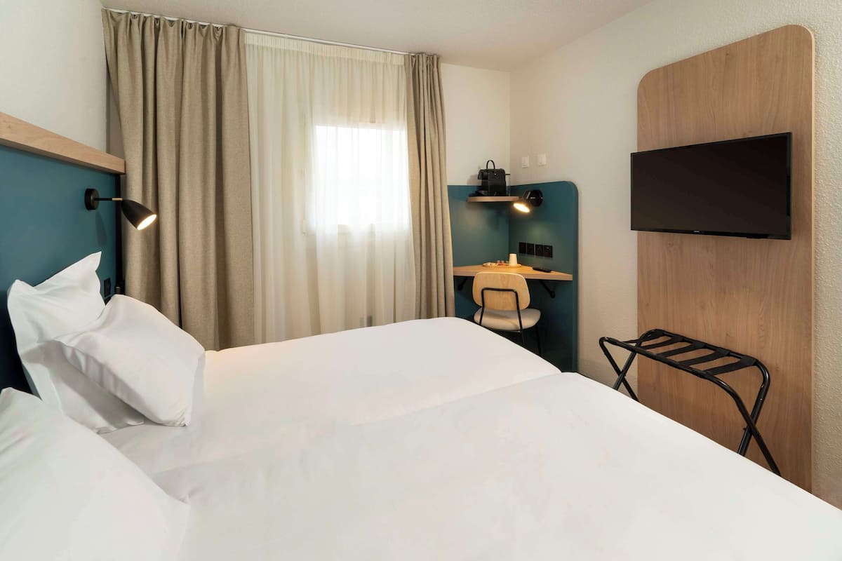 Quarto de hotel com cama, escrivaninha, cadeira, televisão e janela com cortinas.