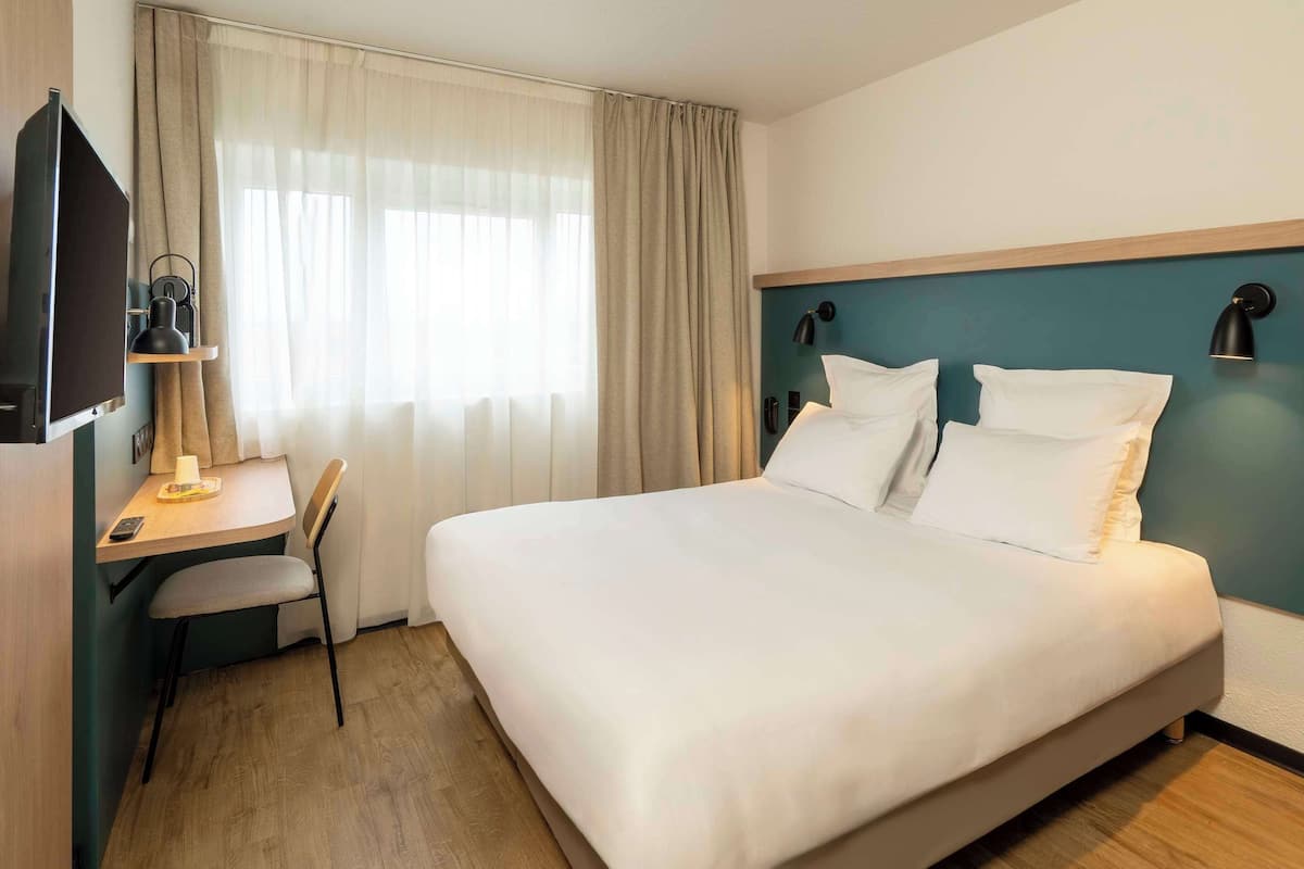 Quarto de hotel com cama, escrivaninha com cadeira, televisão e janela com cortinas.