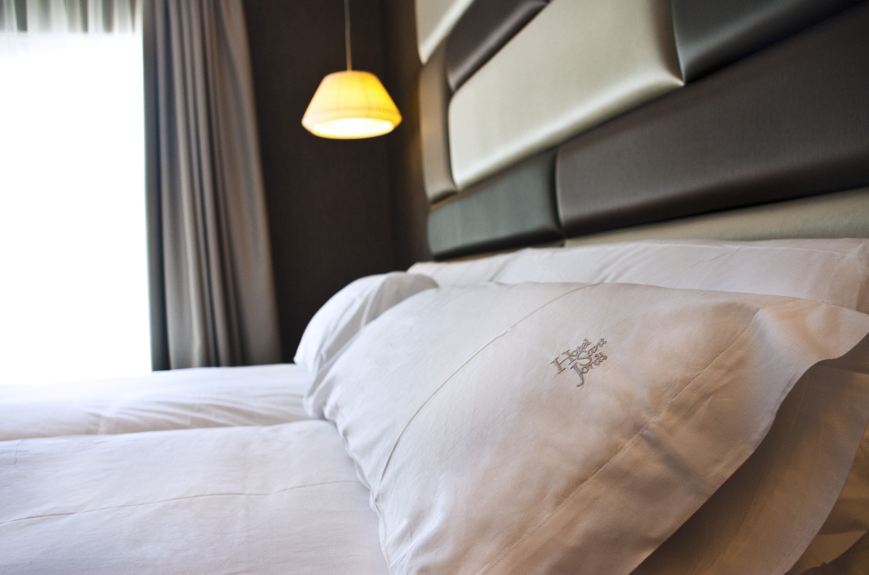 Uma cama bem arrumada com travesseiro ostentando um logotipo, uma luminária pendente e cabeceira.