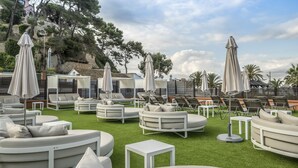 Uma área de lounge no terraço com cadeiras e mesas brancas, cercada por guarda-sóis e vegetação.