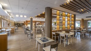Um restaurante espaçoso com piso de madeira, mesas e cadeiras brancas. Janelas amplas permitem entrada de luz natural e vista para o exterior.