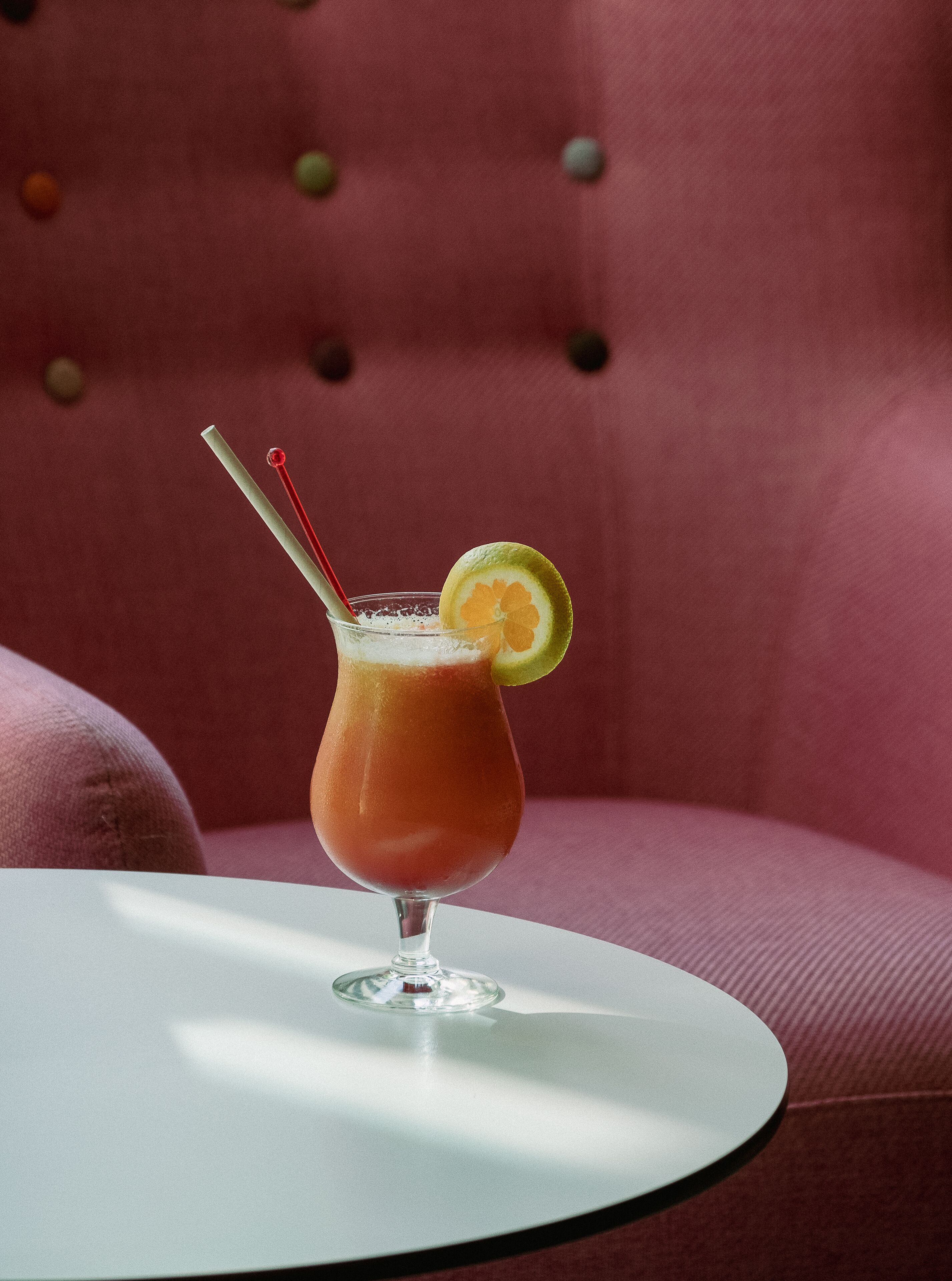 Um cocktail sobre uma mesa branca, com uma cadeira estofada em vermelho ao fundo.