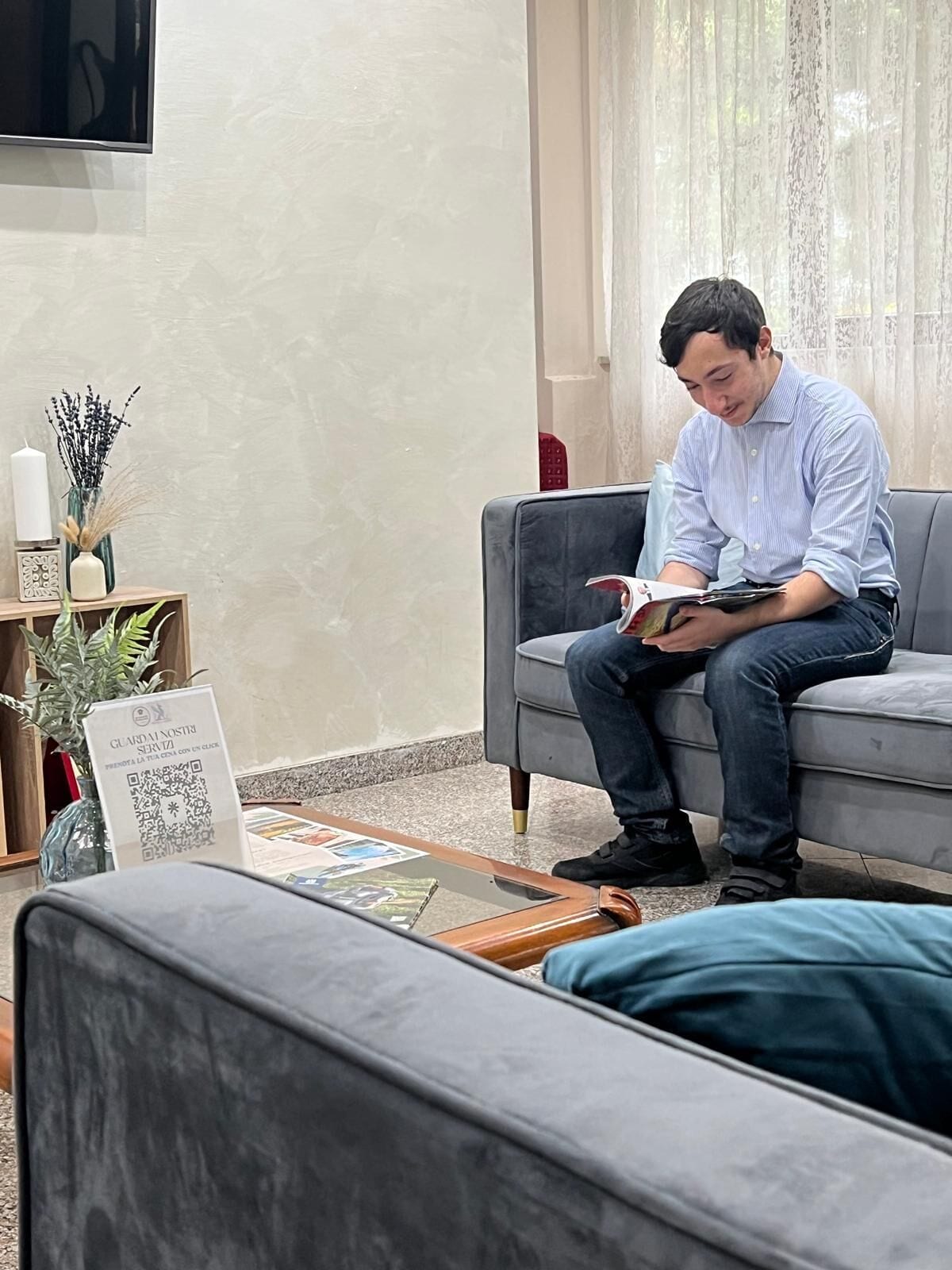 Um homem sentado em um sofá cinza lendo um livro em um quarto com uma mesinha de madeira, um vaso com plantas secas e uma placa emoldurada no chão.