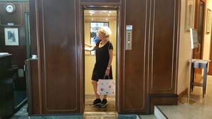 Uma mulher de vestido preto saindo de um elevador, carregando uma bolsa branca.