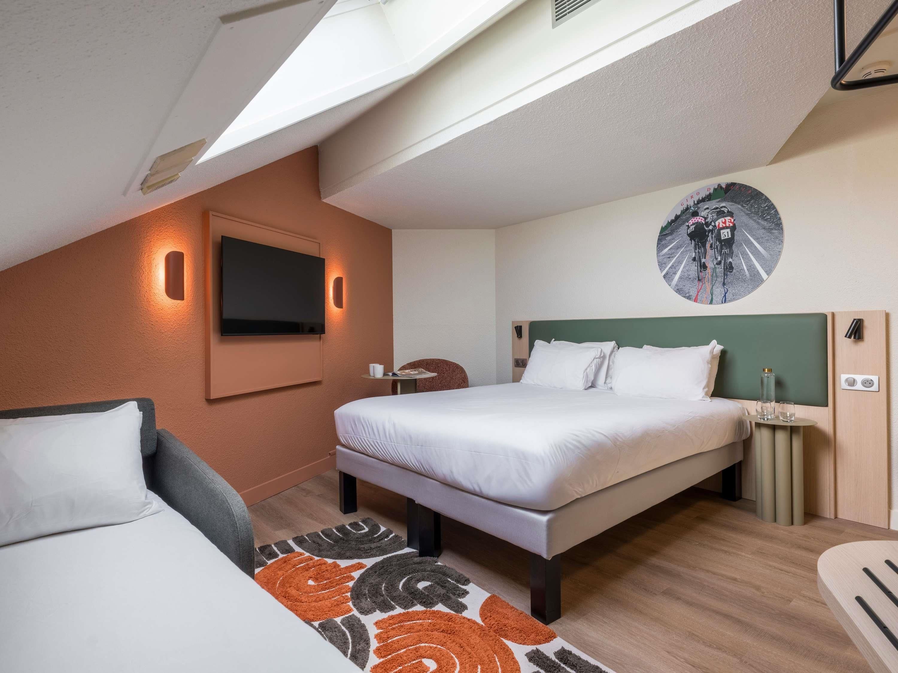 Quarto de hotel com cama, televisão fixada na parede, mesinha com abajur e uma obra de arte circular na parede.