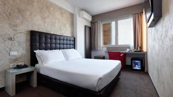 Quarto de hotel com cama, mesa de cabeceira, televisão e pequena área de trabalho.