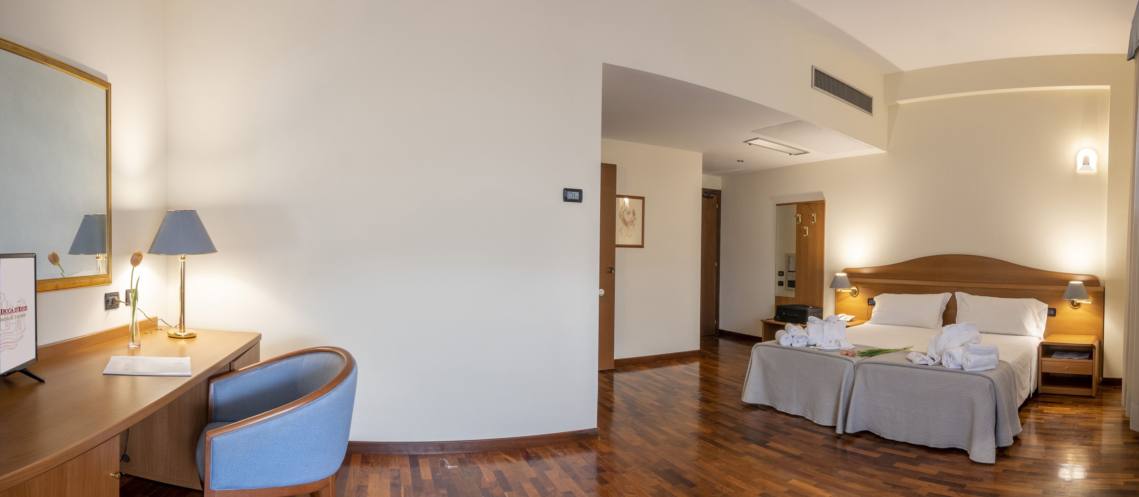 Quarto de hotel com escrivaninha de madeira, duas mesinhas de cabeceira, cama com roupa de cama branca e uma cadeira azul.