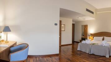 Quarto de hotel com escrivaninha de madeira, duas mesinhas de cabeceira, cama com roupa de cama branca e uma cadeira azul.