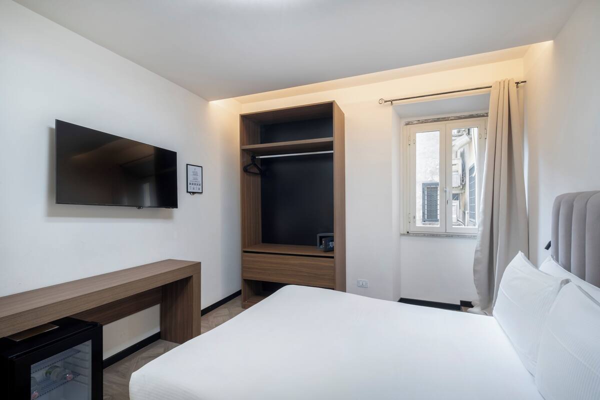 Quarto de hotel com uma cama, televisão fixada na parede, guarda-roupa de madeira, um banco e uma janela com cortinas.