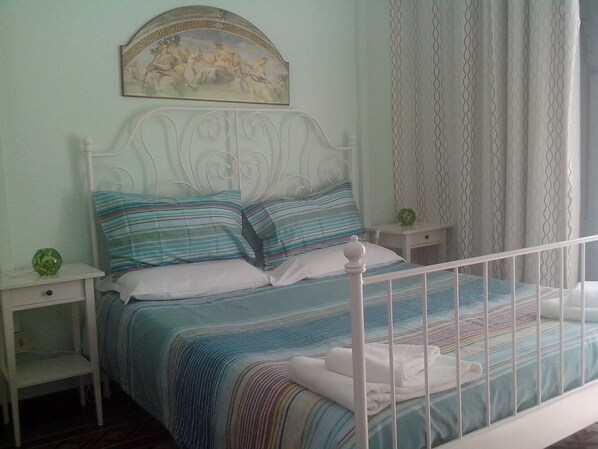 Uma cama com estrutura de metal branca, uma colcha azul e branca listrada e duas almofadas. Há uma pequena mesa de cabeceira com um objeto verde em cima. Um quadro está acima da cama e uma cortina é visível parcialmente à direita.