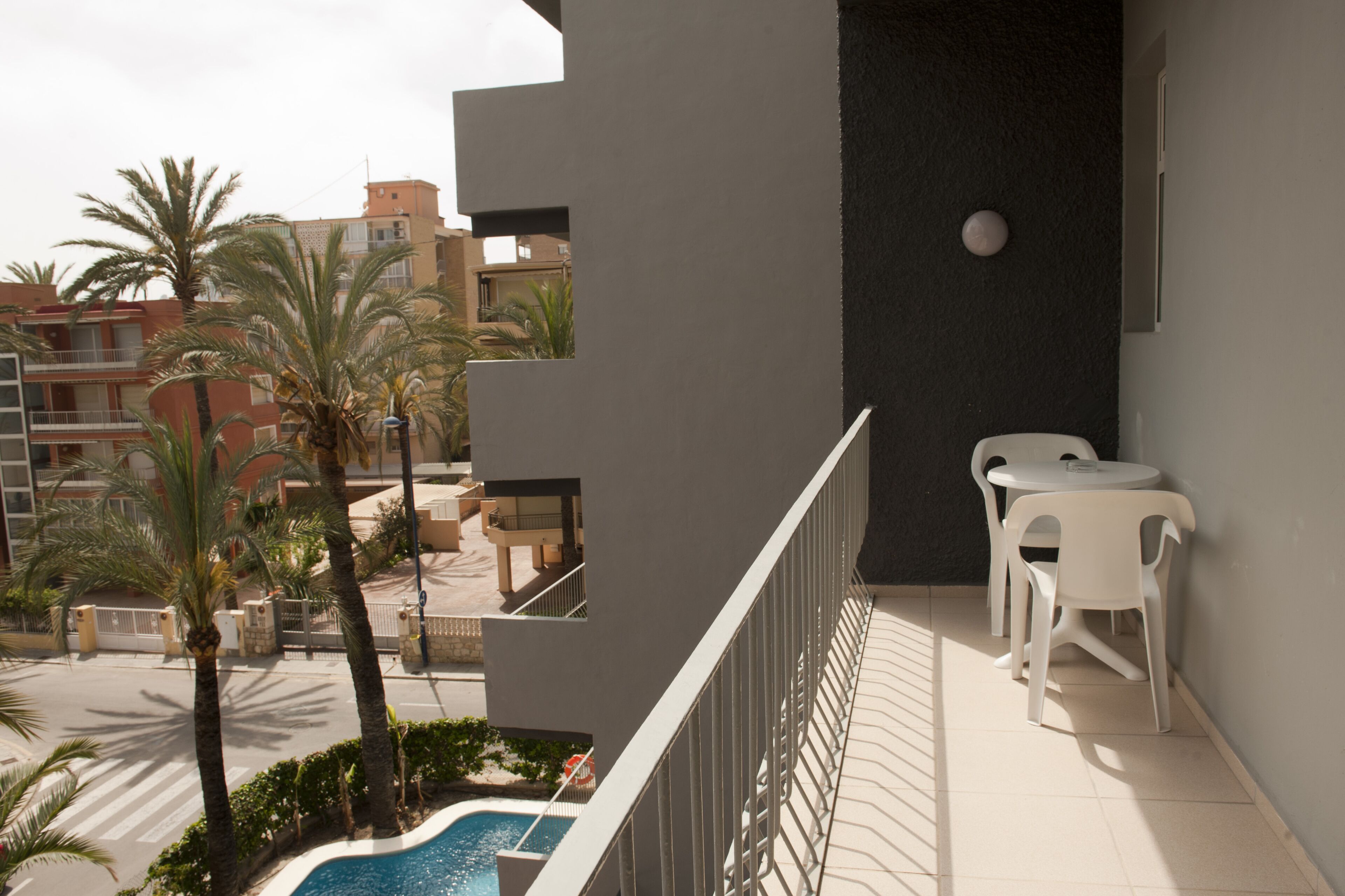 Varanda com piscina, cadeiras brancas e vista para palmeiras e edifícios.