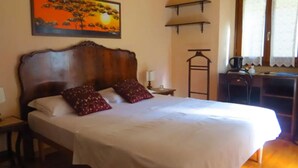 Quarto com cabeceira de madeira, cama com roupa de cama branca e duas almofadas estampadas, mesa de cabeceira com abajur, quadro na parede, escrivaninha com cadeira e janela com cortinas.