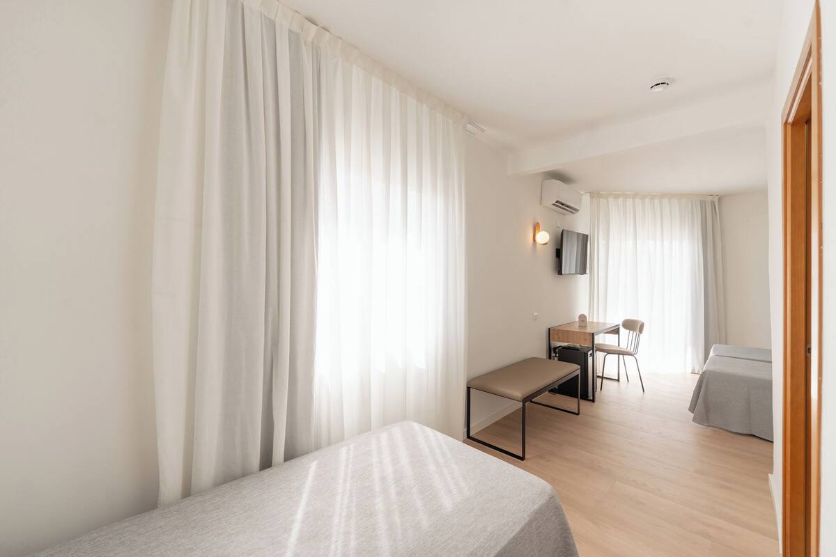 Quarto de hotel com cama, escrivaninha, cadeira, televisão e cama com cobertura cinza.