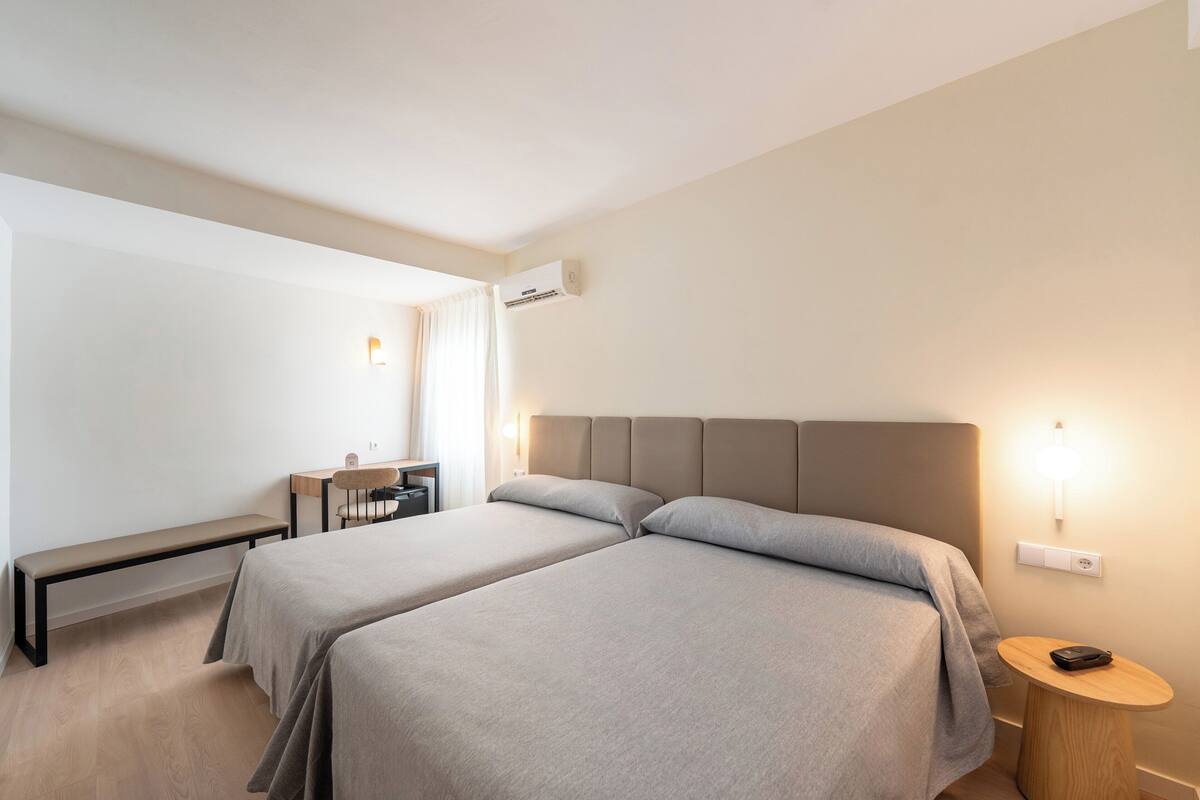 Quarto de hotel com duas camas, uma escrivaninha, um banco e uma pequena mesa com abajur.