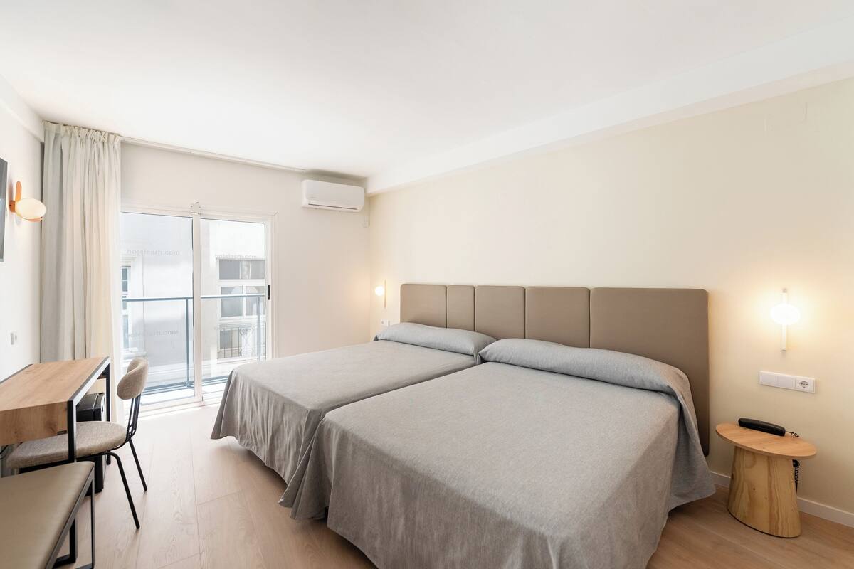 Quarto de hotel com duas camas, uma mesa de escritório, uma cadeira, mesa de cabeceira e vista para o exterior.