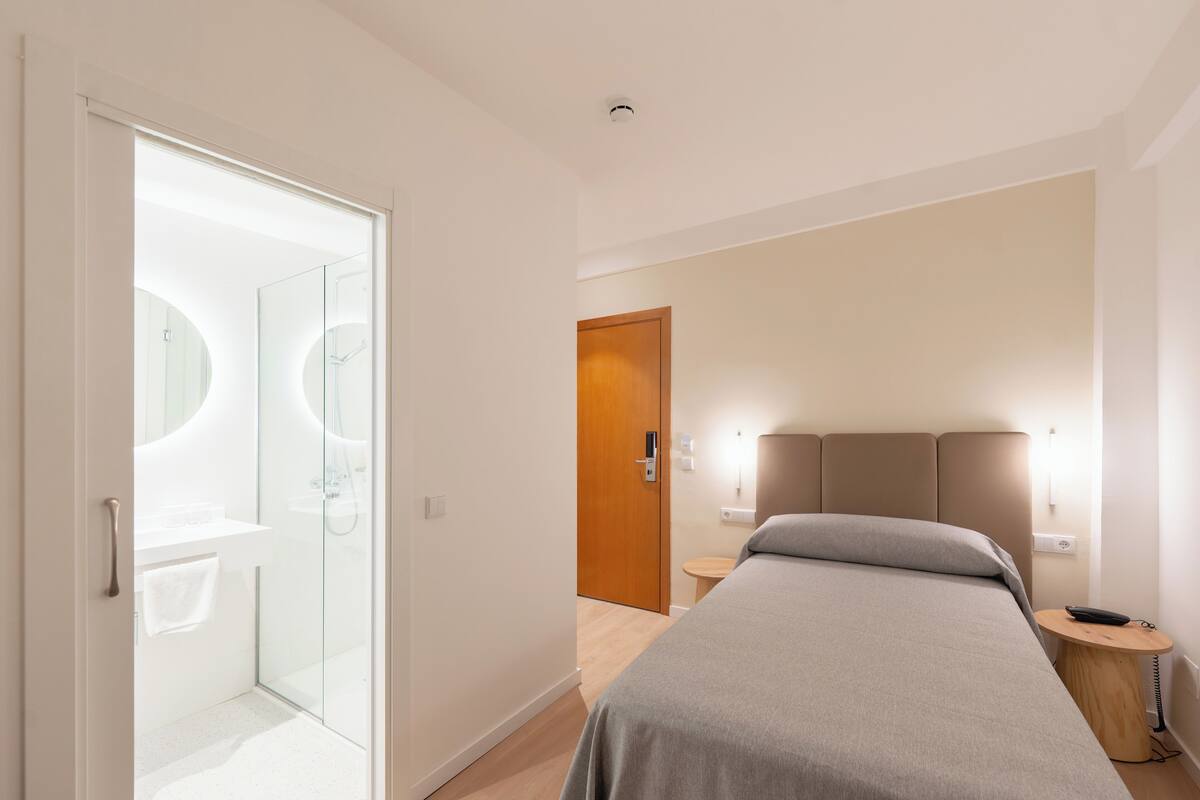 Quarto de hotel moderno com cama, mesa de cabeceira e banheiro com box de vidro.